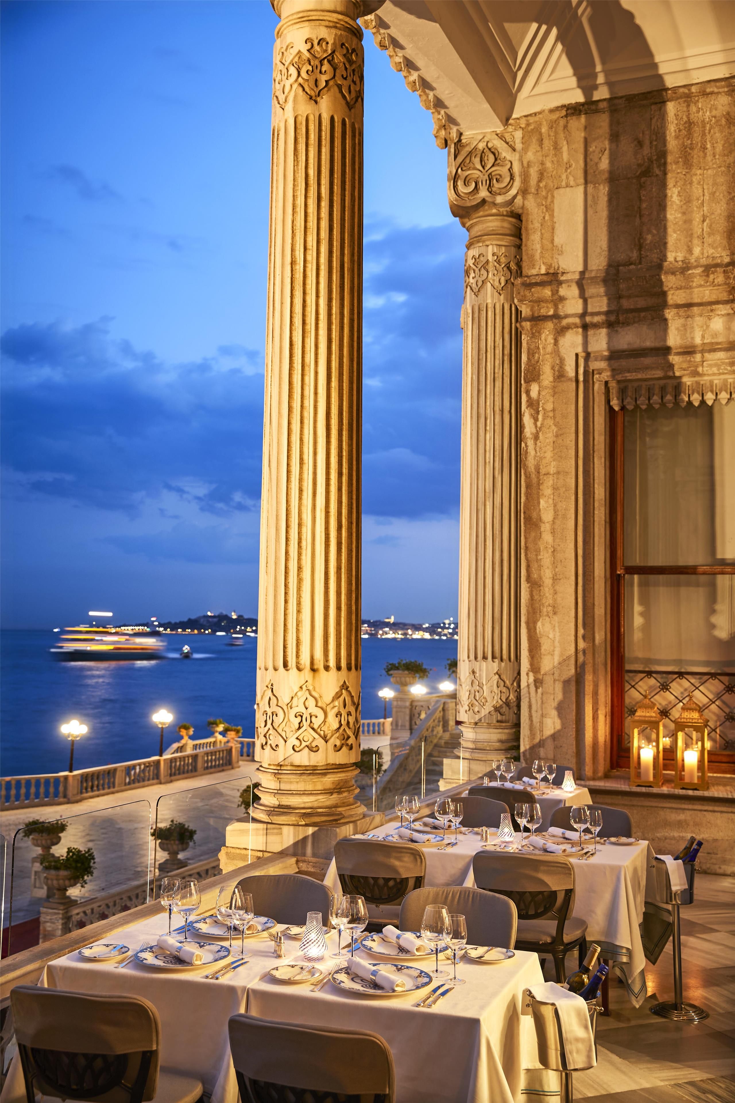 Restaurante com mesas ao ar livre com vista para a água, detalhes em colunas clássicas e vista da cidade.