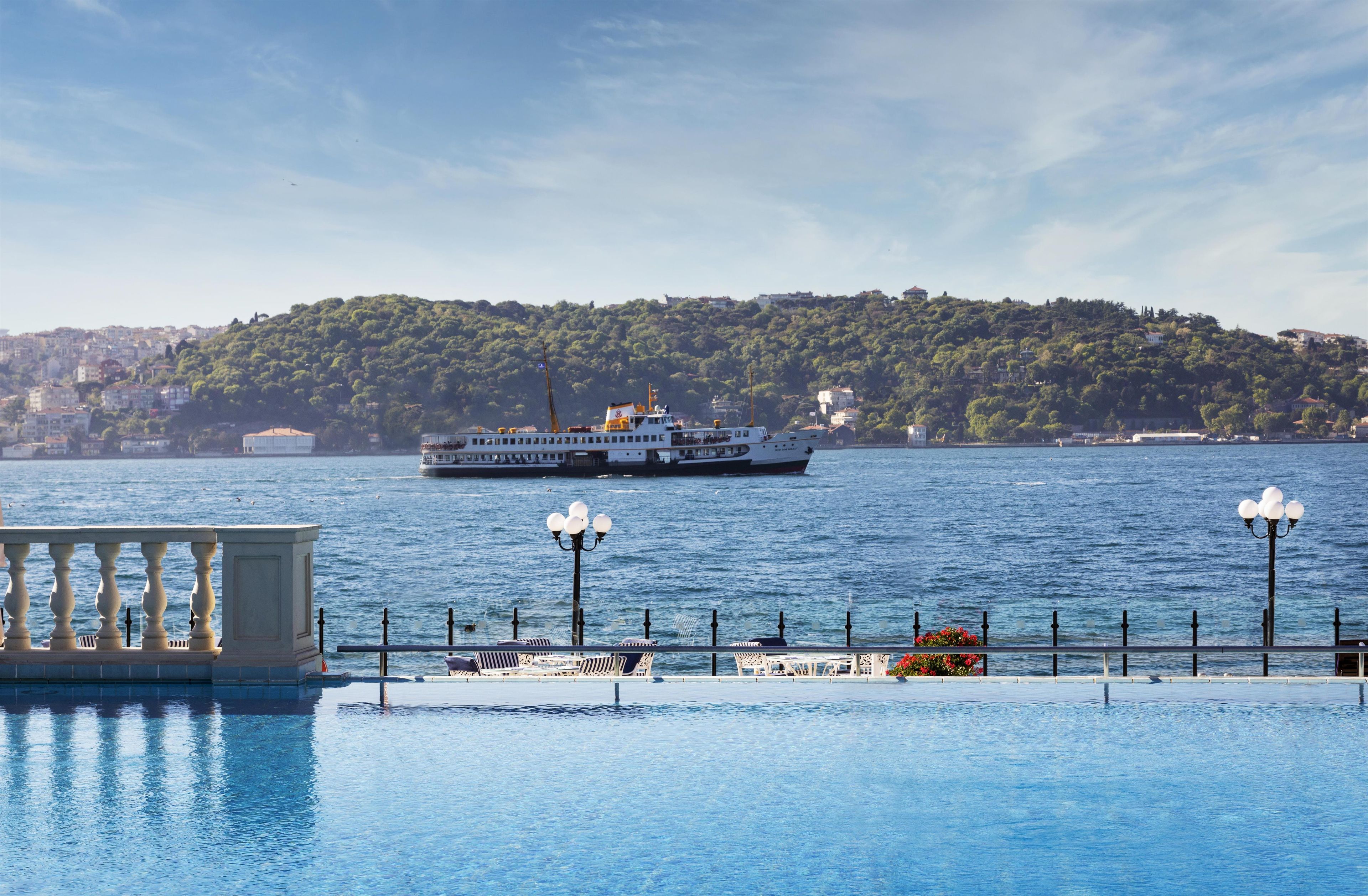 Vista para a orla marítima com um barco de ferry, área da piscina com espreguiçadeiras e um horizonte urbano ao fundo.