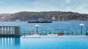 Vista para a orla marítima com um ferry, uma piscina com espreguiçadeiras e uma colina com edifícios ao fundo.