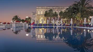 Uma área à beira da piscina com espreguiçadeiras e guarda-sóis, um edifício grande e ricamente decorado ao fundo, e palmeiras.