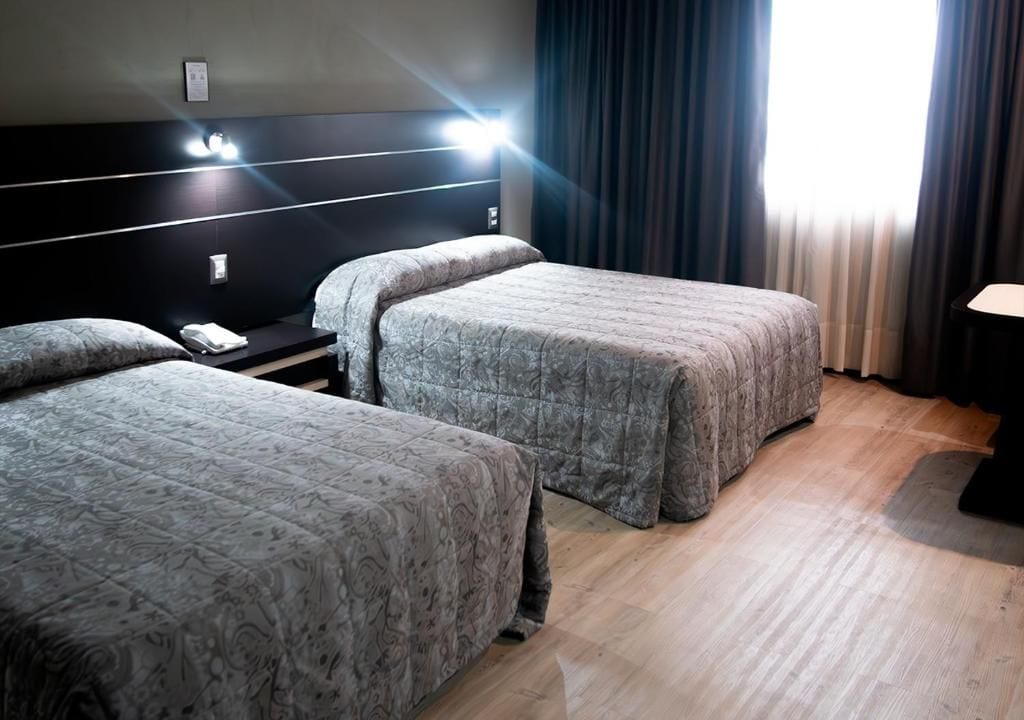 Quarto de hotel com duas camas, uma mesa de cabeceira com abajur e uma janela com cortinas.