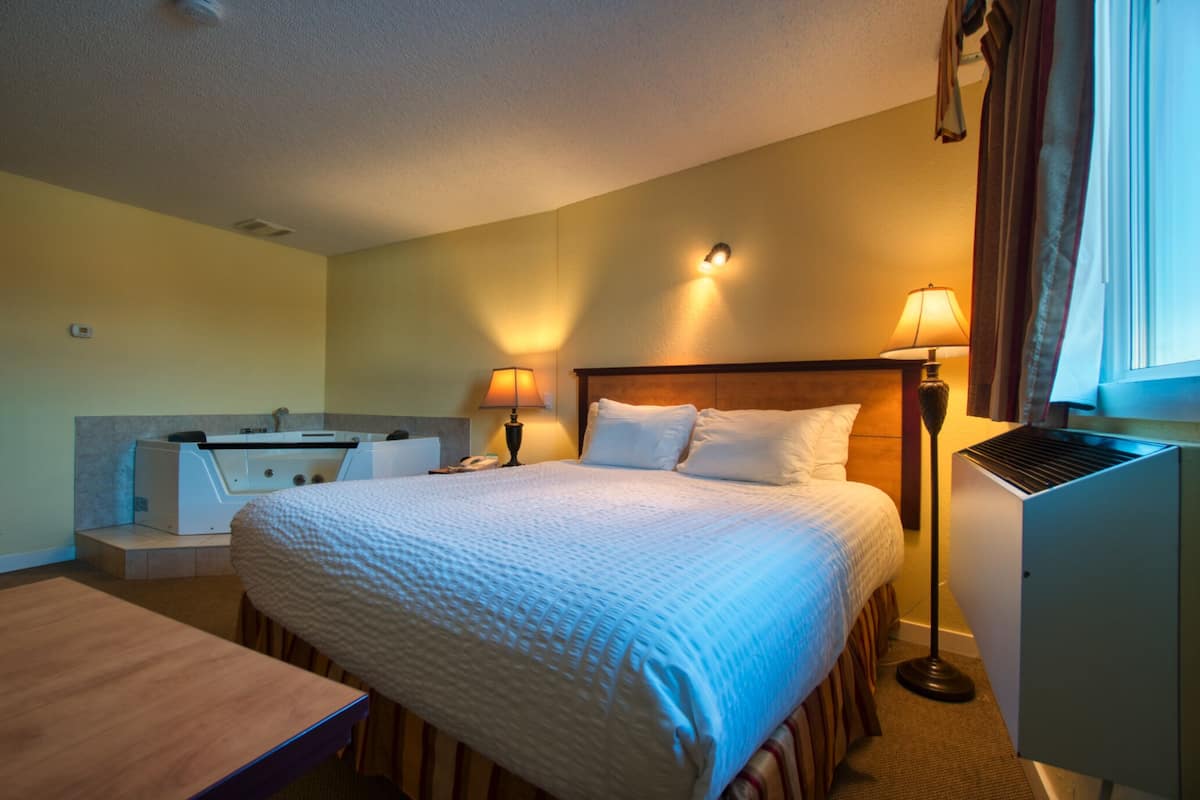 Quarto de hotel com cama, luminárias laterais, uma mesa de trabalho e um pequeno espaço com cozinha.