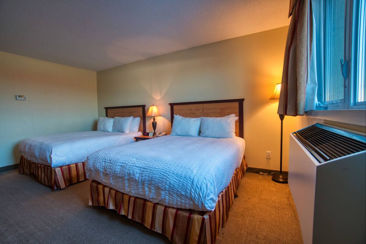 Quarto de hotel com duas camas, uma mesa de cabeceira com abajur, um aquecedor e uma janela com cortinas.