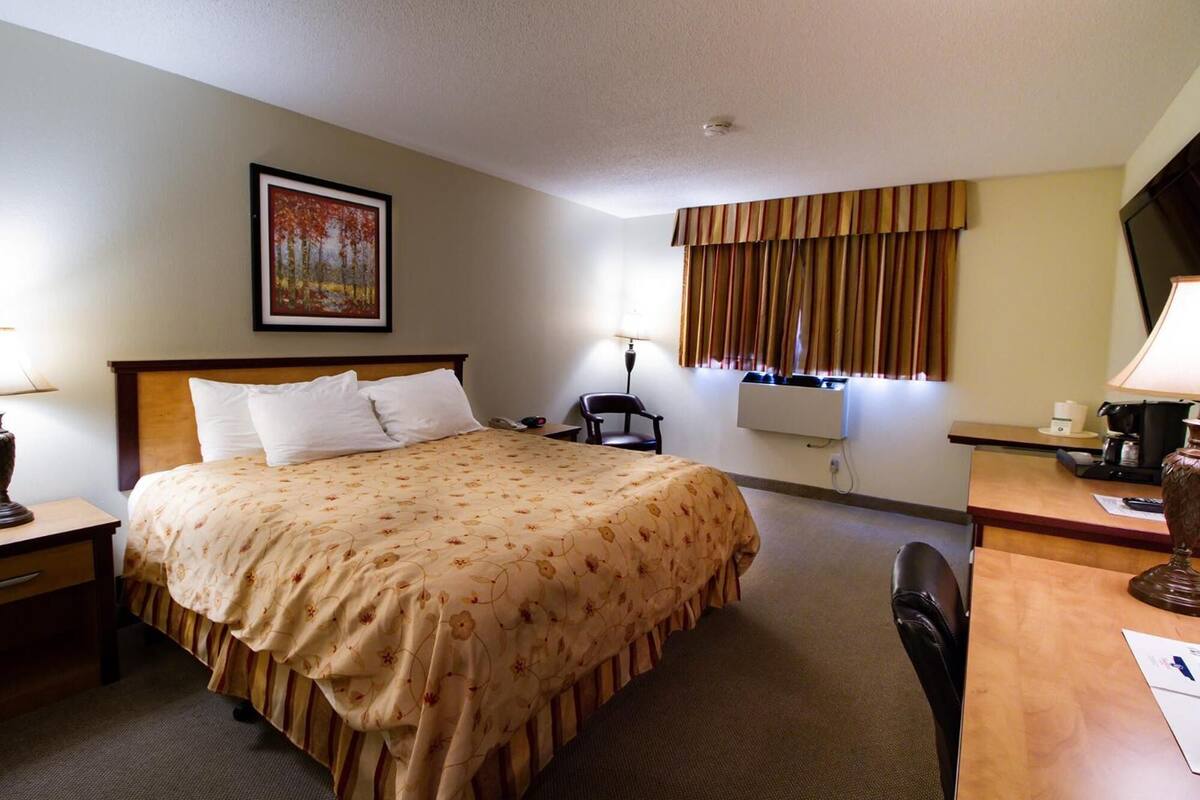 Quarto de hotel com uma cama grande, uma escrivaninha com cadeira, um abajur, um quadro na parede e uma janela com cortinas.