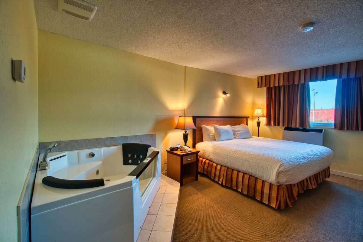 Quarto de hotel com uma cama grande, uma banheira separada, mesa de cabeceira com abajur e janela com cortinas.