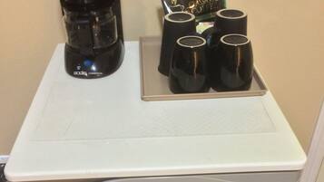 Uma pequena cozinha com cafeteira, quatro chávenas e um pacote de café no balcão.