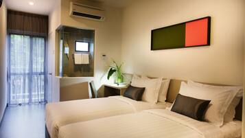 Quarto de hotel com duas camas, uma secretária com cadeira, um vaso de planta e uma janela grande com cortinas.