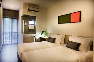 Quarto de hotel com duas camas, uma secretária com cadeira, um vaso de planta e uma janela grande com cortinas.