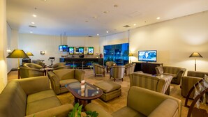 Um hall de hotel com sofás, poltronas, uma mesa de centro, um bar e uma televisão exibindo um noticiário.