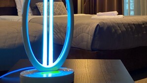Quarto de hotel com uma cama, abajur e luminária circular sobre uma mesa.