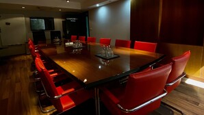 Uma sala de conferências com uma mesa comprida, cadeiras vermelhas e uma tela de televisão.