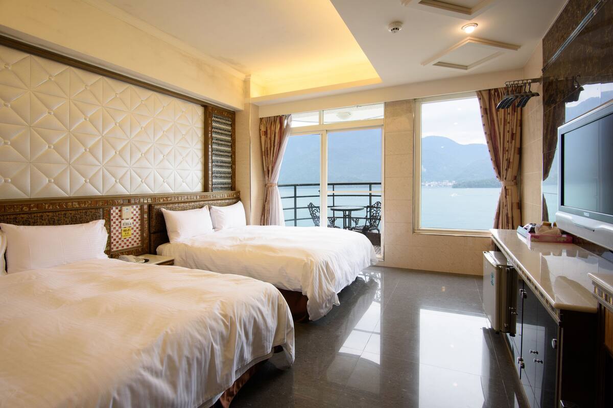 Quarto de hotel com duas camas, uma janela grande com vista para o mar e uma televisão ao lado direito.