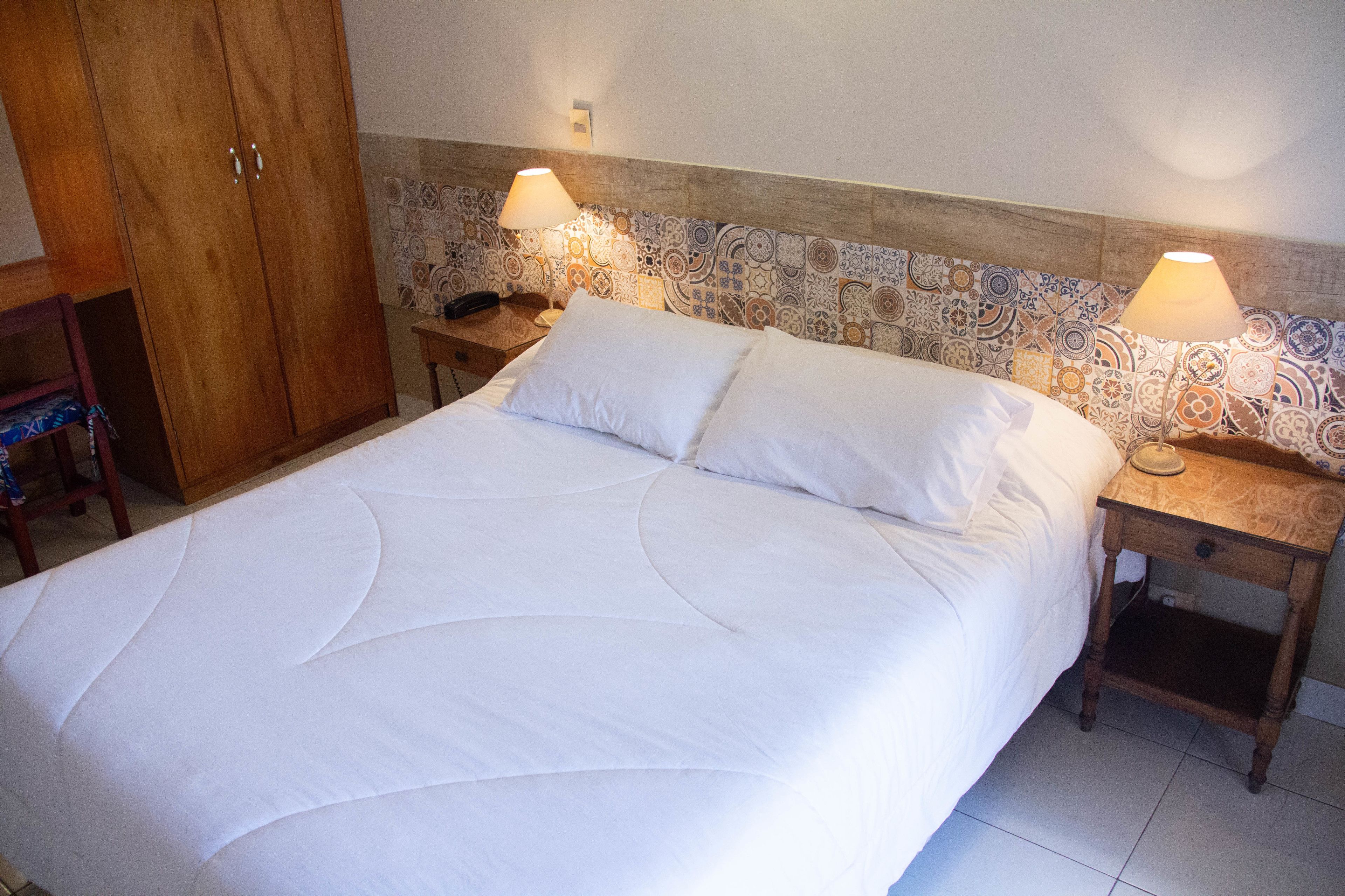Uma cama bem arrumada com lençóis brancos, duas mesas de cabeceira com abajures, um guarda-roupa de madeira e um revestimento de azulejos estampados.