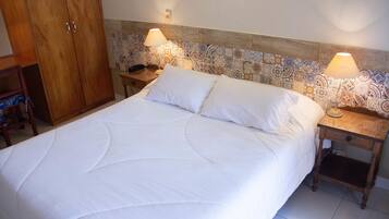Uma cama bem arrumada com lençóis brancos, duas mesas de cabeceira com abajures, um guarda-roupa de madeira e um revestimento de azulejos estampados.