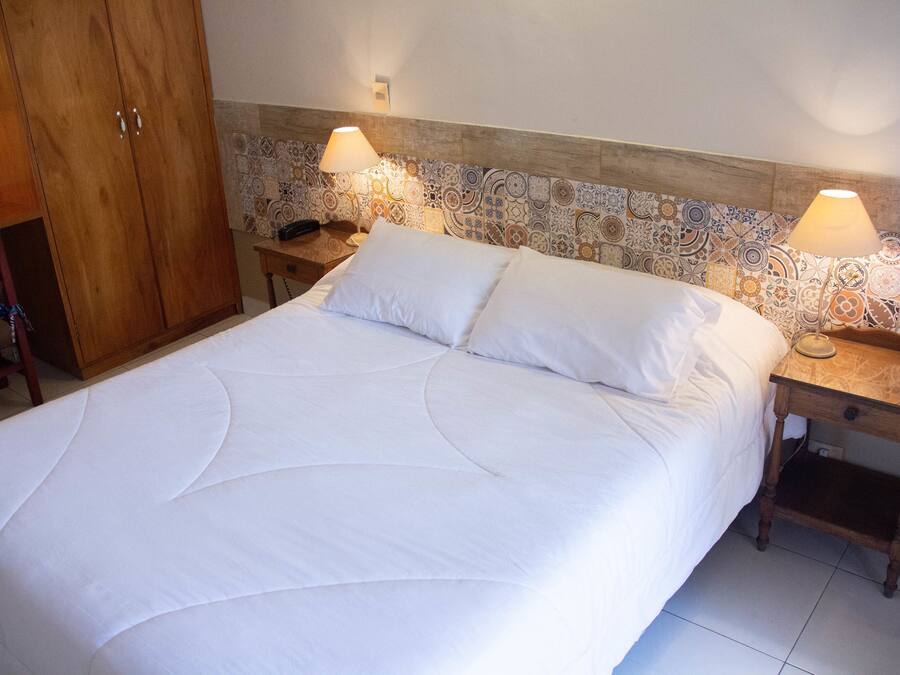 Uma cama bem arrumada com lençóis brancos, duas mesas de cabeceira com abajures, um guarda-roupa de madeira e um revestimento de azulejos estampados.