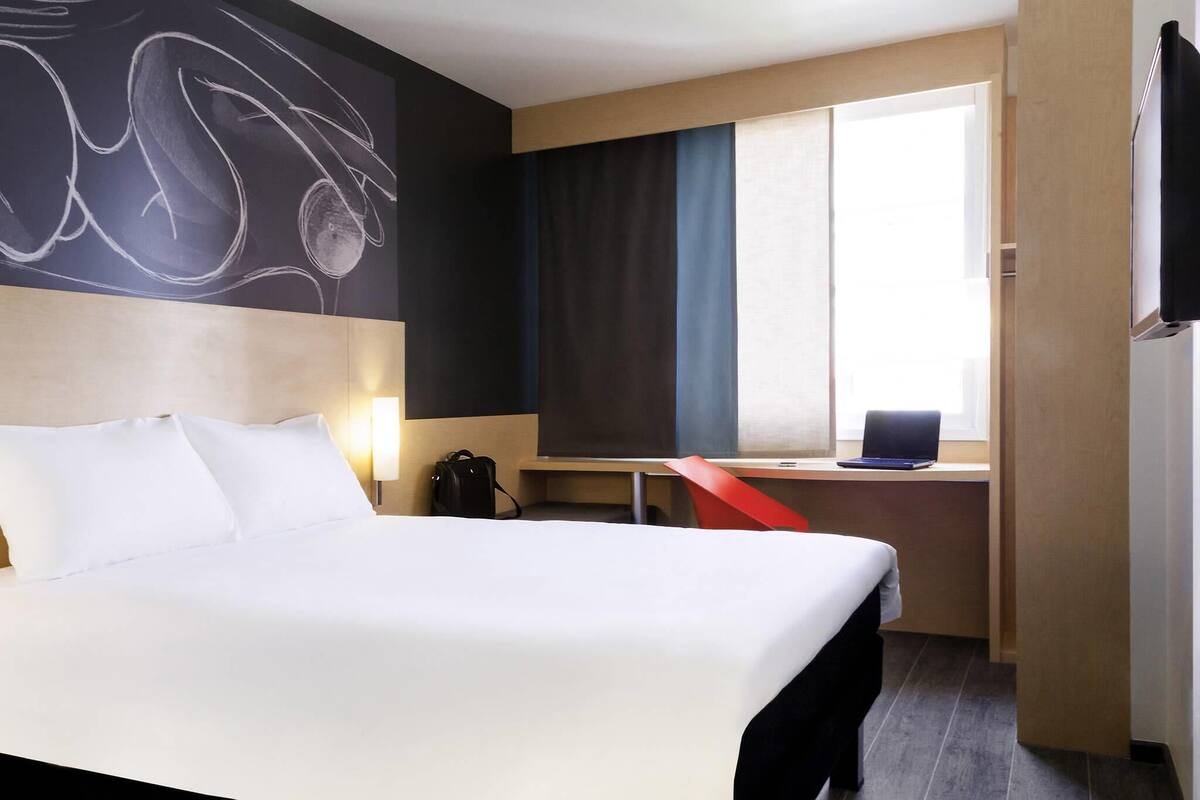 Quarto de hotel com uma cama grande, uma mesa com um laptop, uma janela com cortinas e um painel decorativo na parede.
