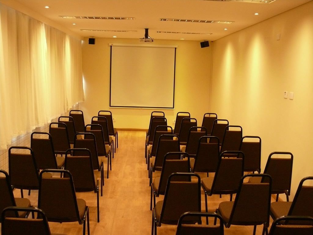 Sala de conferências com fileiras de cadeiras pretas voltadas para uma grande tela de projeção.