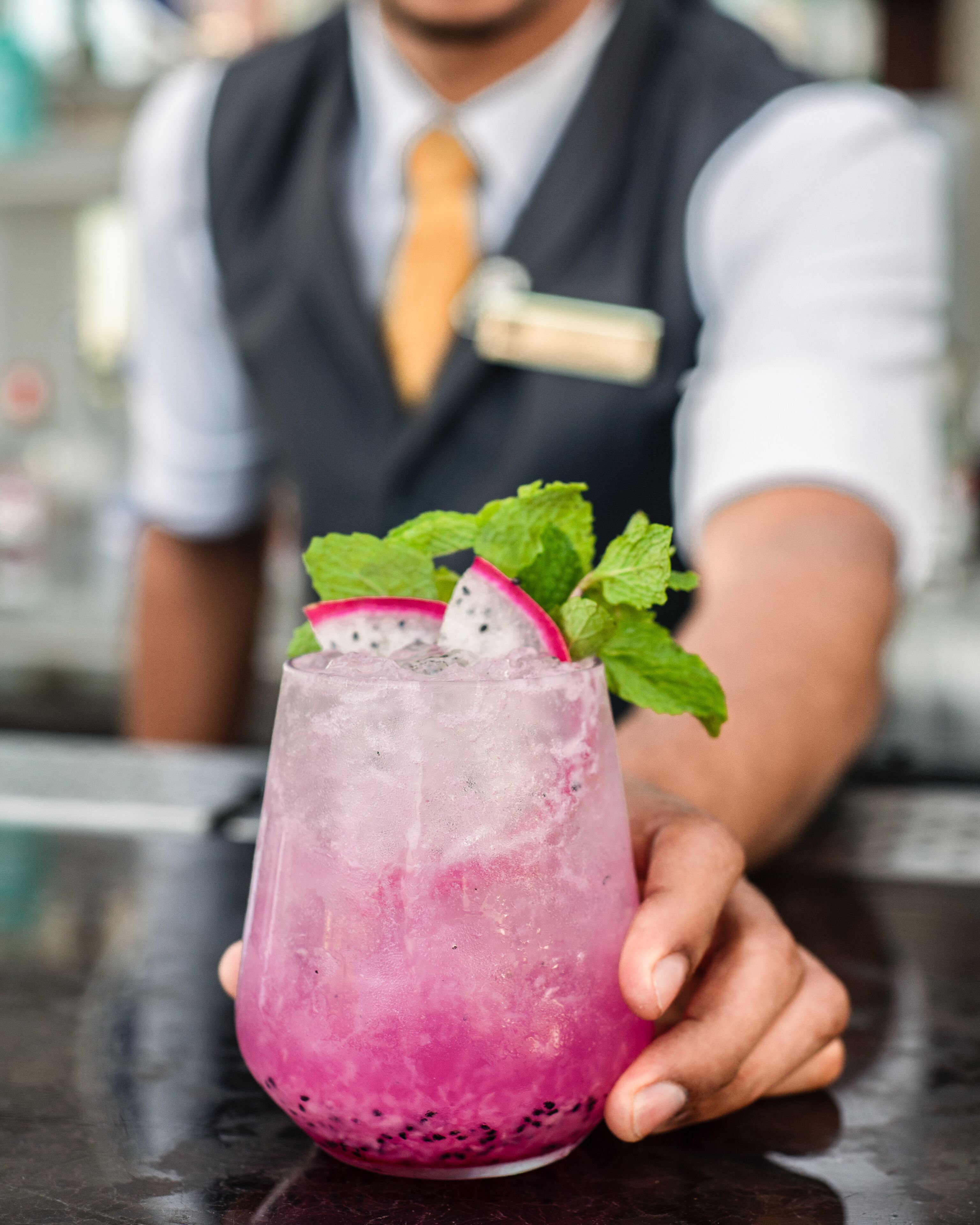 Um bartender segurando um copo com bebida rosa, decorado com folhas de hortelã e fatias de pitaya.