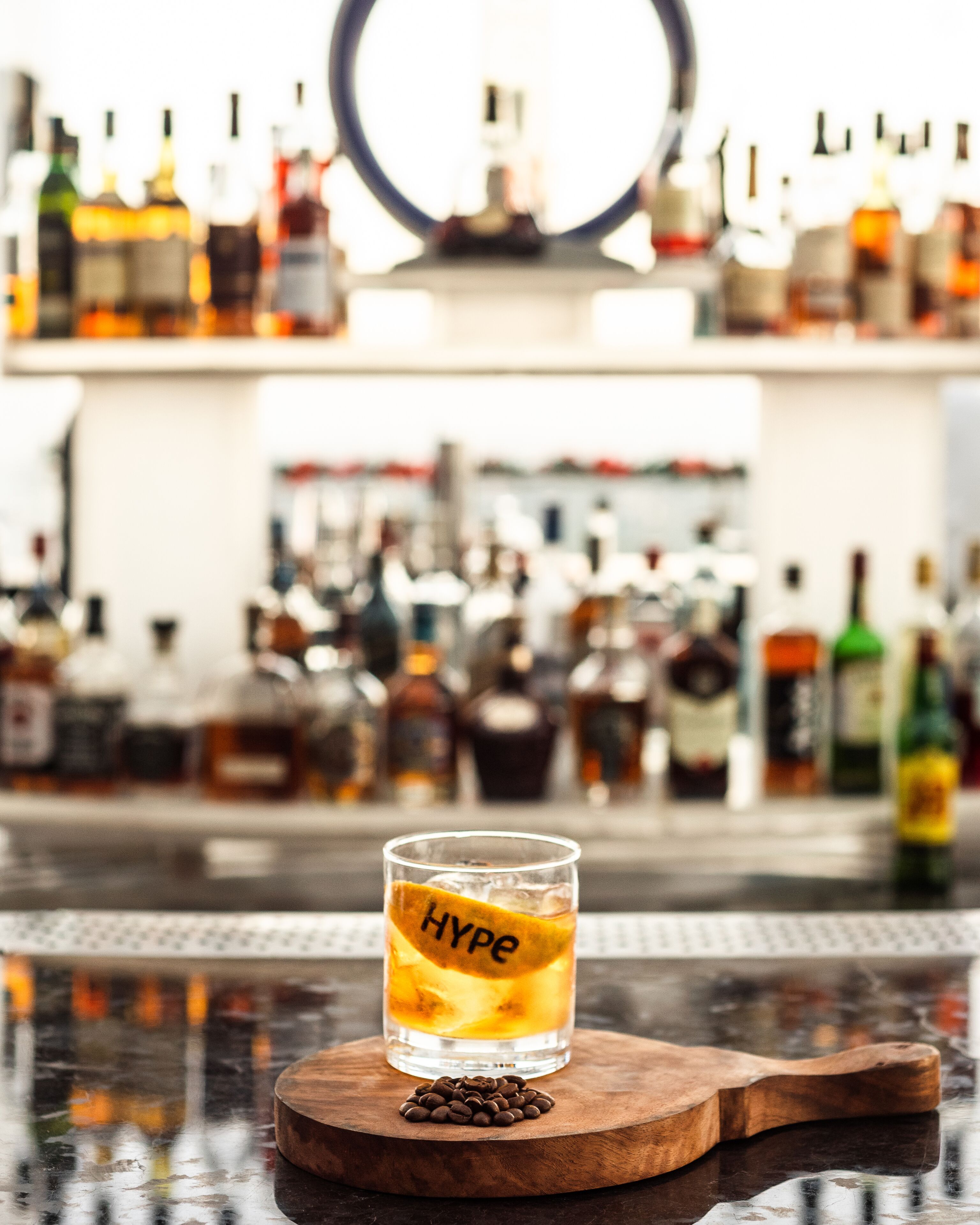 Um bar com um copo identificado com a palavra 'Hype' sobre um apoio de madeira, rodeado por diversas garrafas e um espelho circular ao fundo.