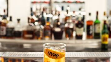 Um bar com um copo identificado com a palavra 'Hype' sobre um apoio de madeira, rodeado por diversas garrafas e um espelho circular ao fundo.