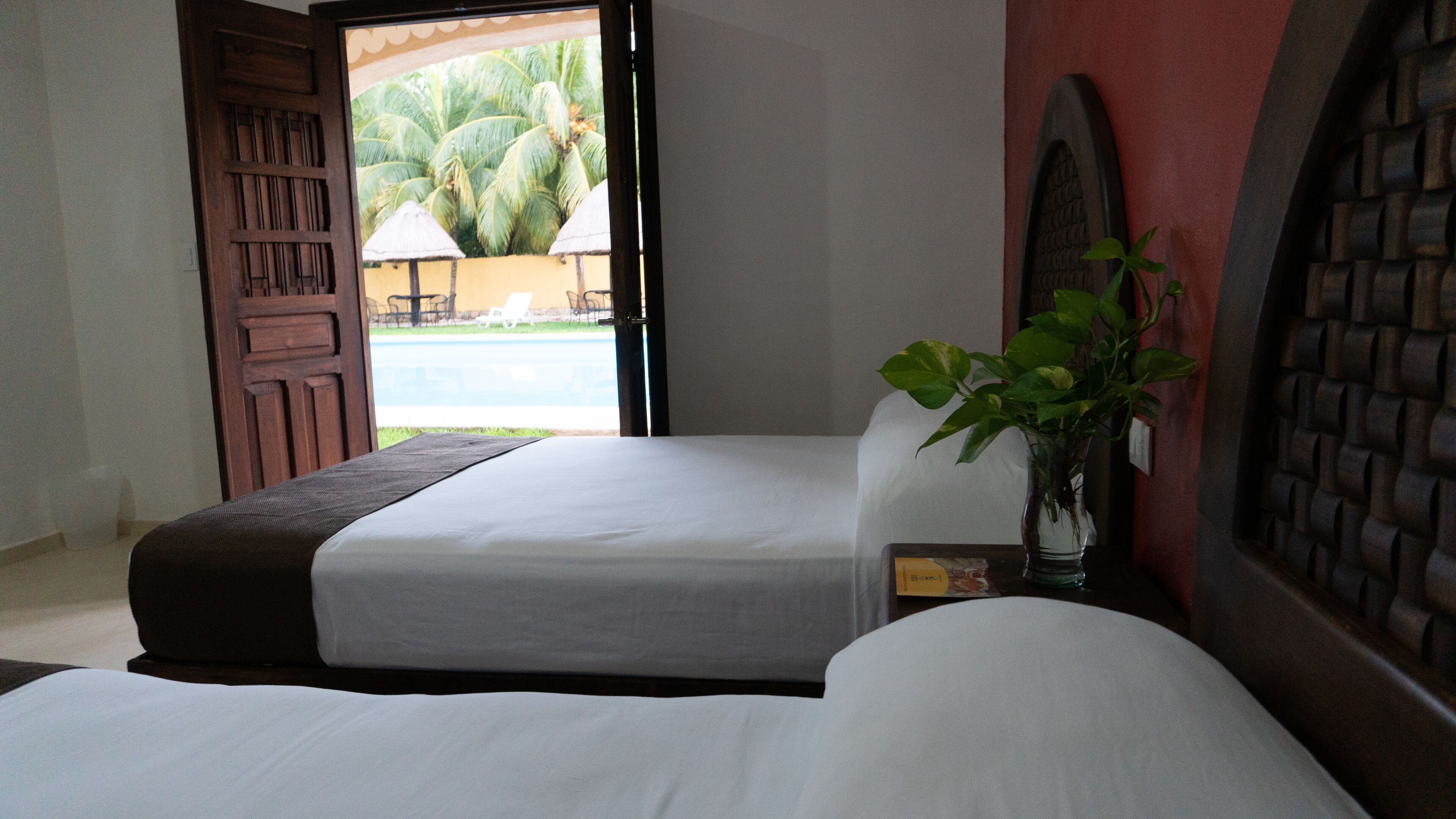 Quarto de hotel com duas camas, porta de madeira, vista para piscina e palmeiras, e um vaso de planta.