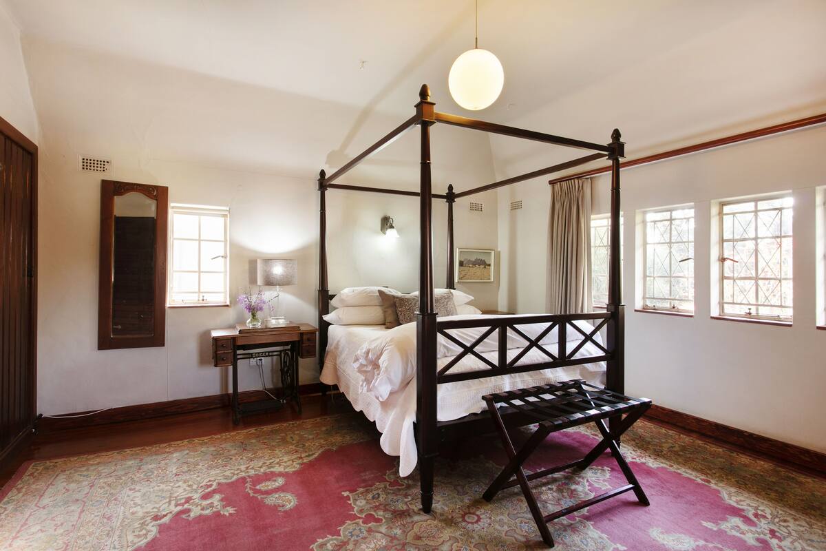 Uma cama com dossel, um banquinho, uma mesa de cabeceira, um espelho e uma janela com cortinas.
