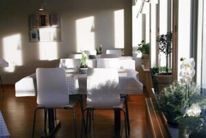Uma sala de jantar com mesa posta para quatro pessoas, rodeada por cadeiras brancas, com plantas em vasos e vista para o exterior.