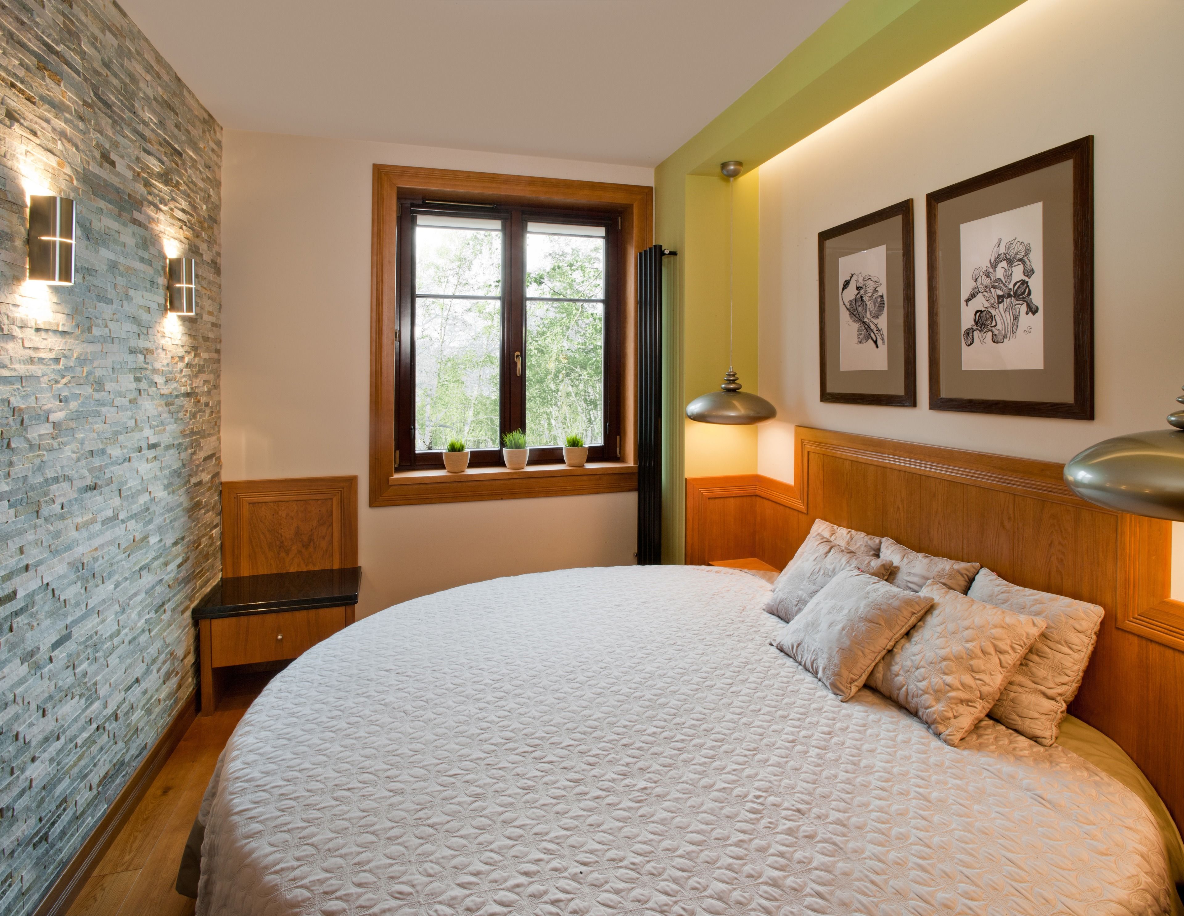 Quarto com uma cama redonda grande, parede de pedra, janela com plantas e quadros nas paredes.
