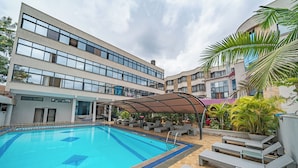 Piscina com espreguiçadeiras e área de estar coberta, cercada por edifícios com janelas amplas e uma palmeira.