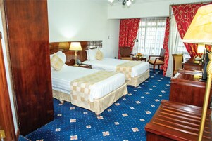 Quarto de hotel com duas camas, uma escrivaninha, uma cadeira e uma mesa pequena com uma luminária.