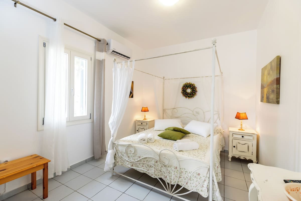 Uma cama de dossel com colcha florida, duas mesas de cabeceira com abajures, um banco de madeira e um ar-condicionado fixado na parede.