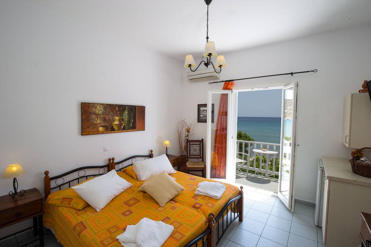 Um quarto com cama, mesa de cabeceira, escrivaninha, cadeira, um quadro e vista para o mar através de uma porta aberta.