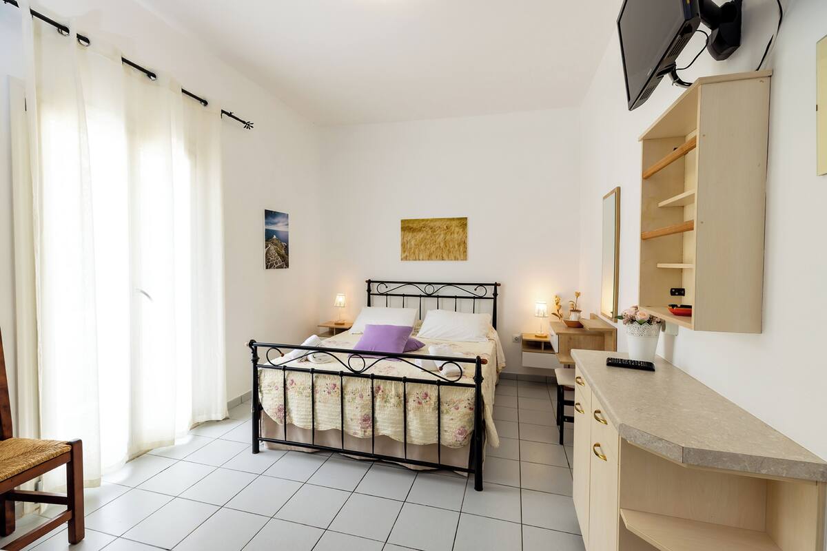 Quarto com cama, mesas de cabeceira, escrivaninha e televisão na parede.