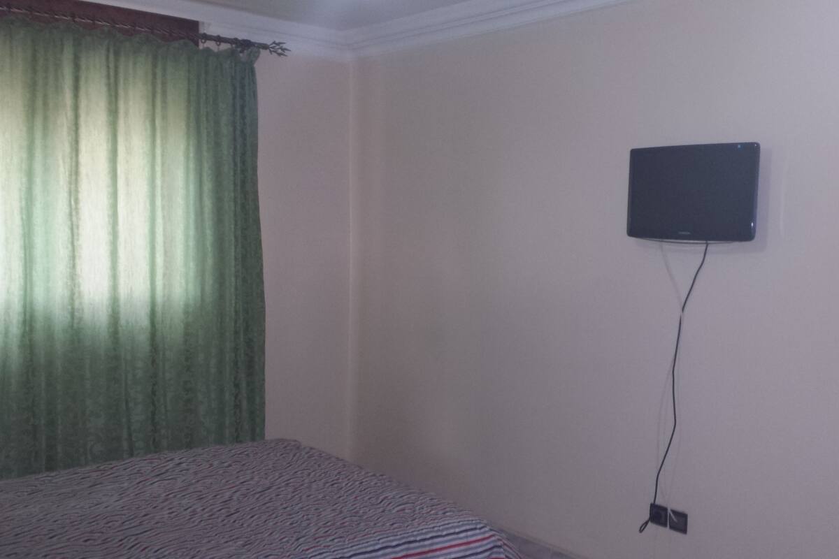 Quarto com cama, cortina verde e televisão na parede.