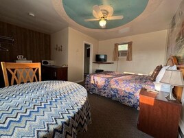 Quarto com cama, cadeira, escrivaninha, televisão e ventoinha no teto.