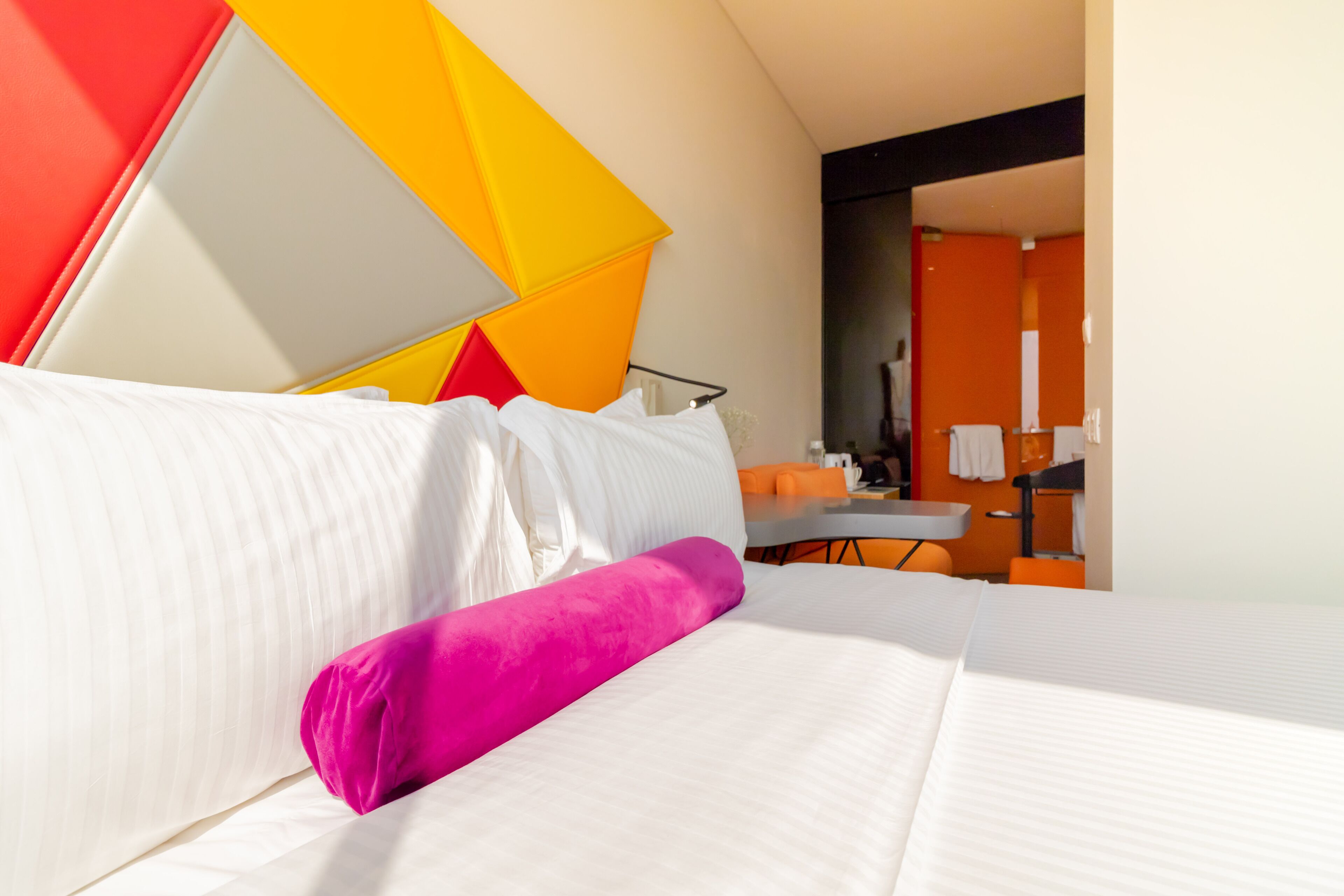 Quarto de hotel com uma cama, almofadas, uma mesa e um banheiro visível por uma porta aberta.