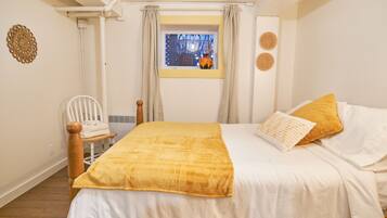 Quarto com cama, janela com cortinas, uma cadeira e um objeto decorativo na parede.