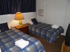 Quarto de hotel com duas camas, uma mesa de cabeceira com abajur, uma pequena prateleira e um guarda-roupa.