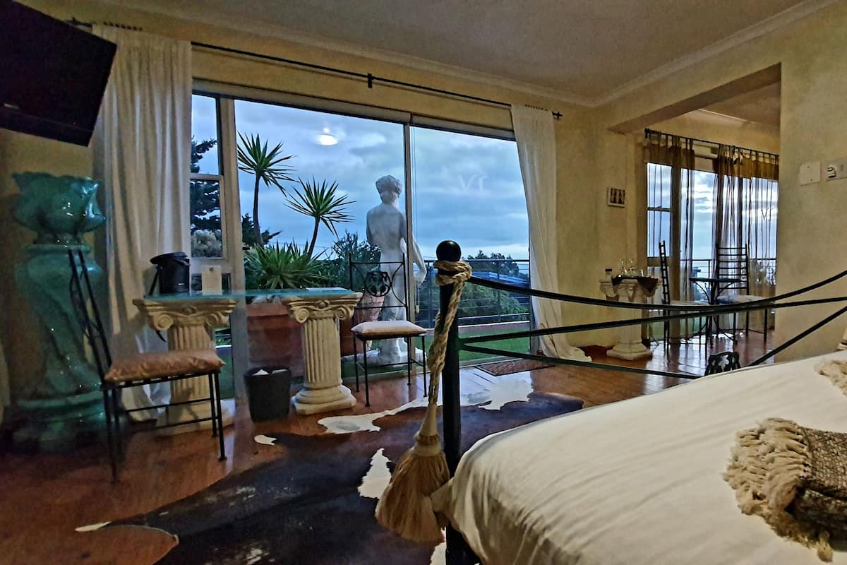 Quarto de hotel com uma cama grande, vista para o mar, uma estátua e um espaço de jantar também com vista.