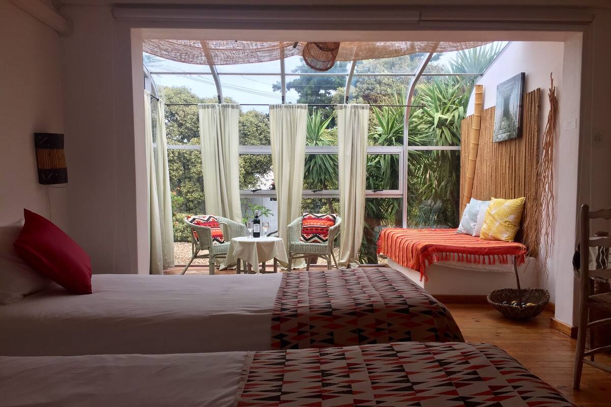 Quarto com duas camas, uma mesa de jantar com cadeiras e vista para o verde através de amplas janelas.