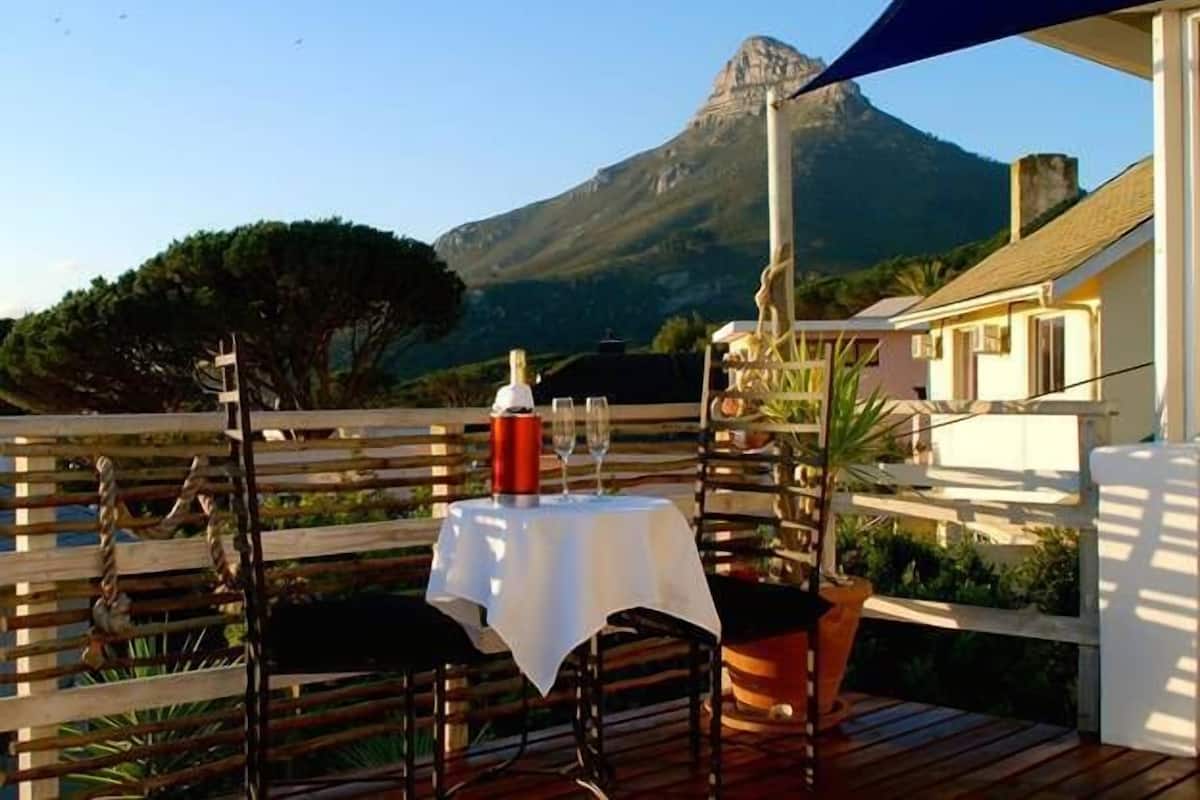 Mesa com toalha branca, duas cadeiras e uma garrafa vermelha em um deck com vista para a montanha.