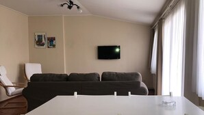 Sala de estar com uma mesa branca e quatro cadeiras verdes, um sofá cinza, uma televisão na parede e um lustre no teto.