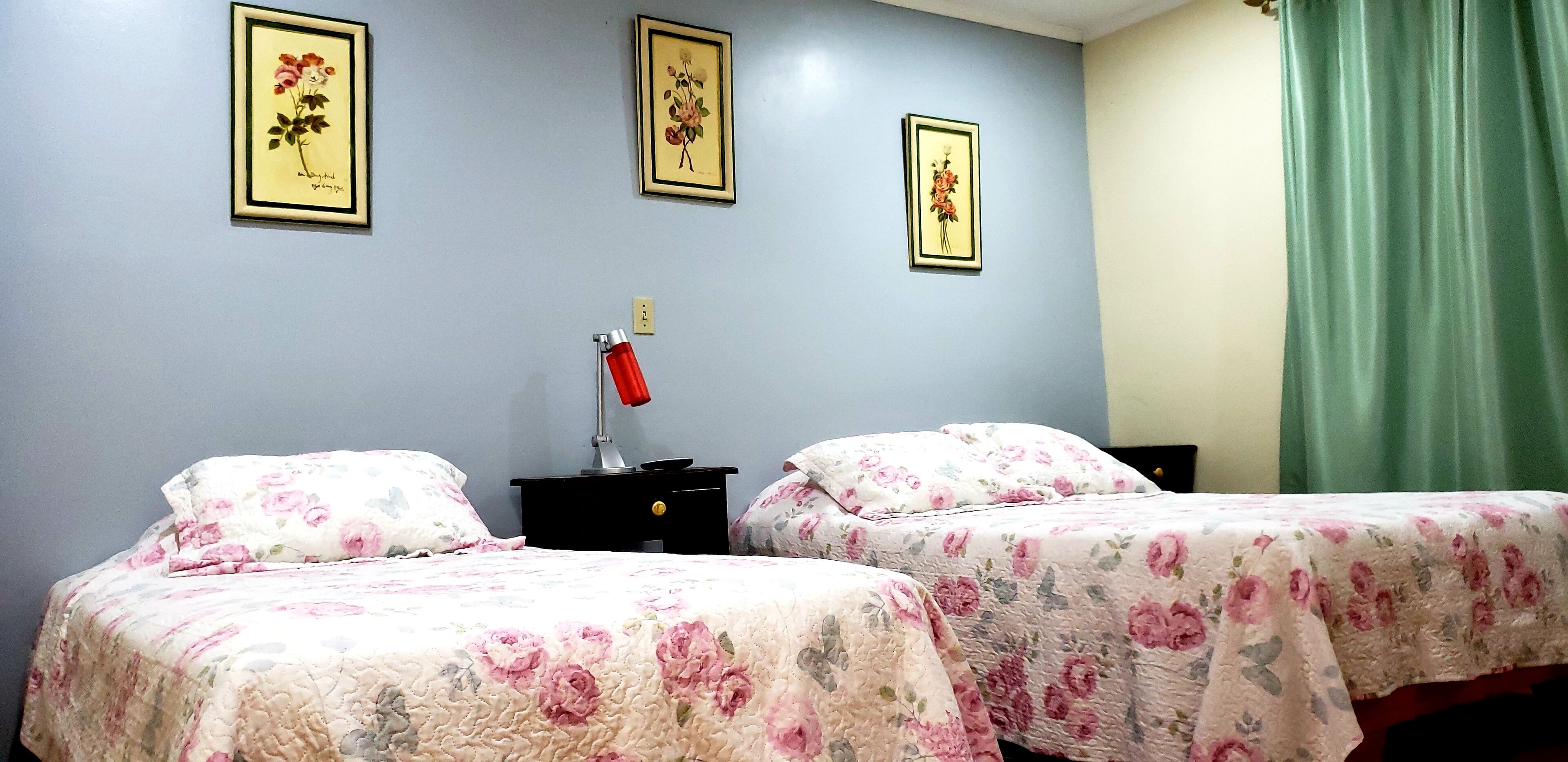 Quarto com duas camas, cada uma com roupa de cama florida, uma mesinha de cabeceira com abajur vermelho e três quadros florais na parede.