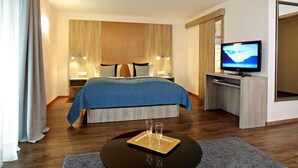 Quarto de hotel com cama, mesas de cabeceira, televisão e uma pequena mesa com copos.
