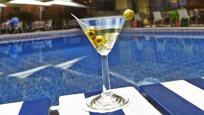 Vista clara de uma piscina com uma taça de martini em uma espreguiçadeira, um edifício de hotel ao fundo e guarda-sóis.