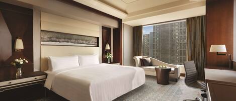 Quarto de hotel com uma cama grande, uma escrivaninha, uma cadeira, uma mesinha com flores e vista para edifícios altos.