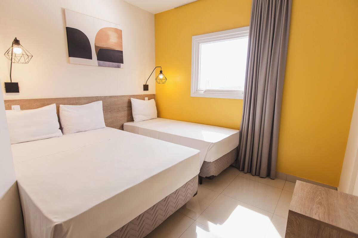 Quarto de hotel com duas camas, uma janela com cortinas e luminárias fixadas na parede.