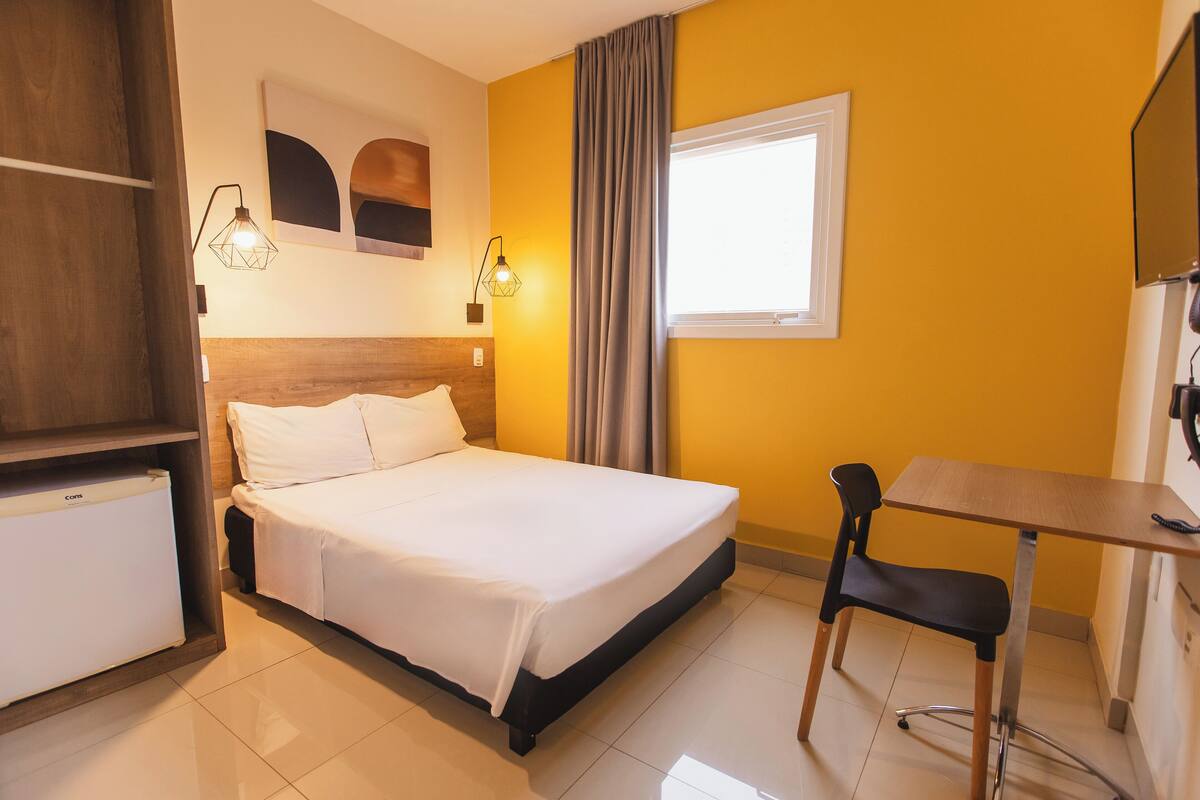 Quarto de hotel com cama, escrivaninha, cadeira, televisão e janela com cortinas.