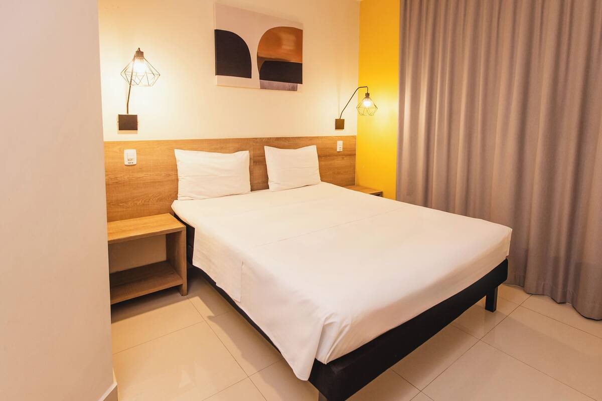 Uma cama bem arrumada com lençóis brancos, cabeceira de madeira e duas luminárias laterais.