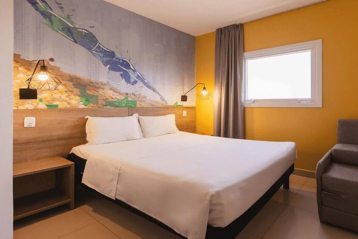 Quarto de hotel com uma cama grande, área de estar com uma poltrona, painel decorativo na parede e janela com cortinas.